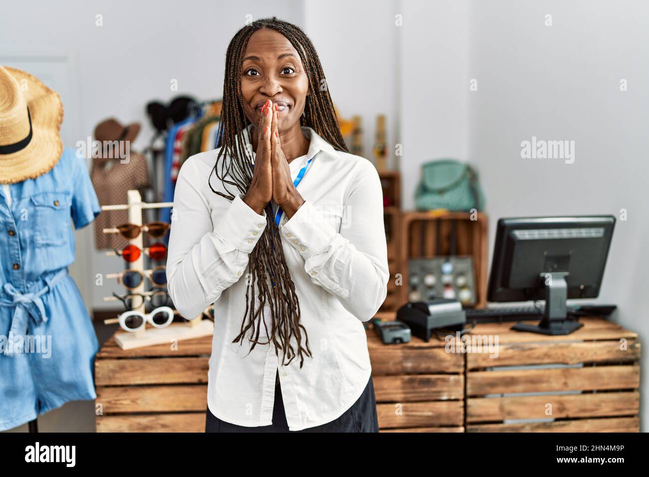 Black woman with braids working as manager at retail boutique praying with hands together asking ...
