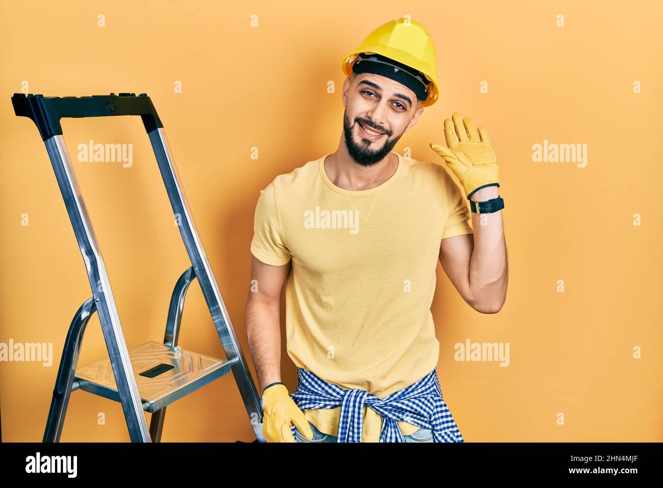 Handsome man with beard by construction stairs wearing hardhat waiving ...