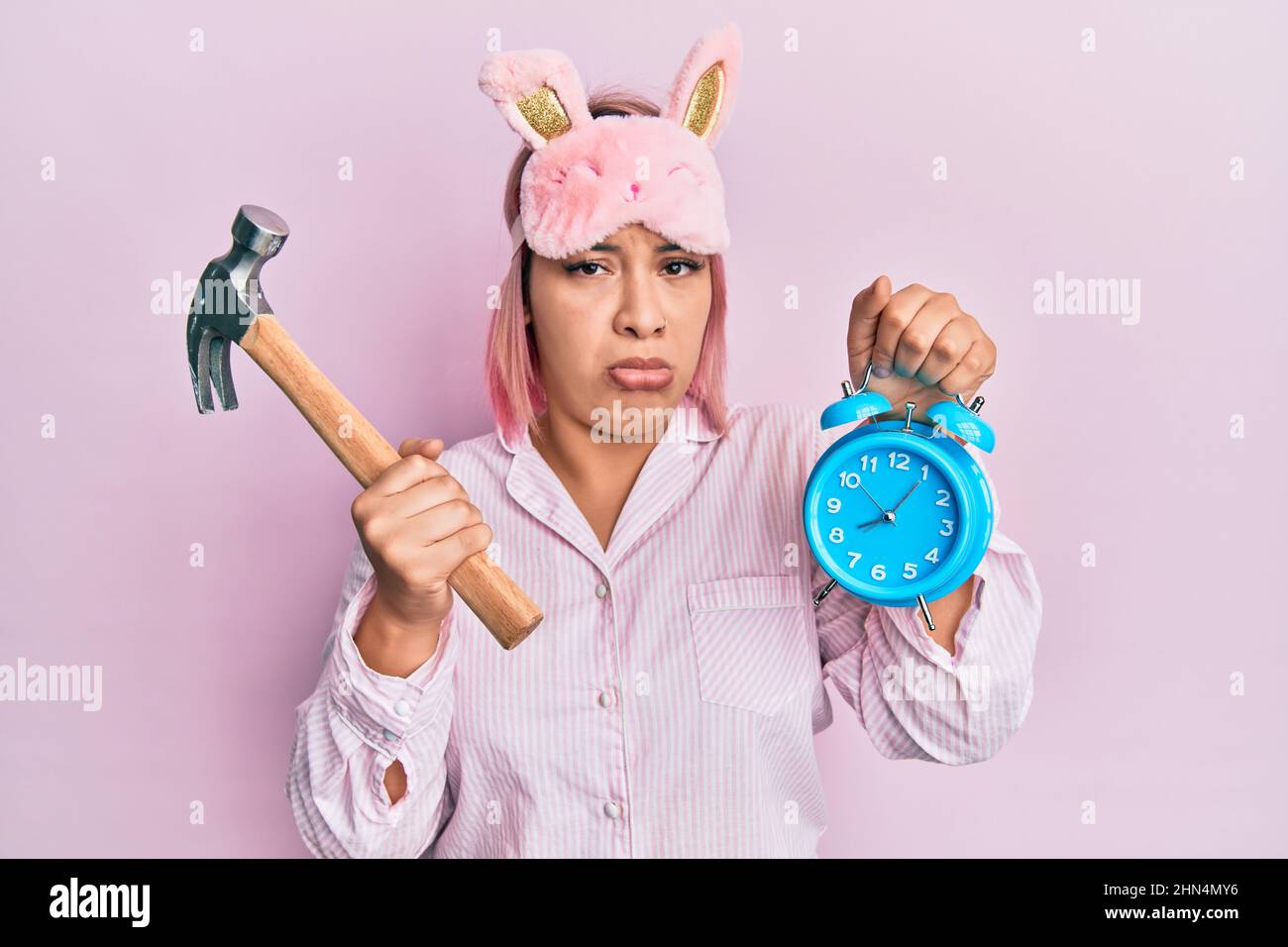 Hispanic woman with pink hair wearing pajama holding hammer and alarm ...