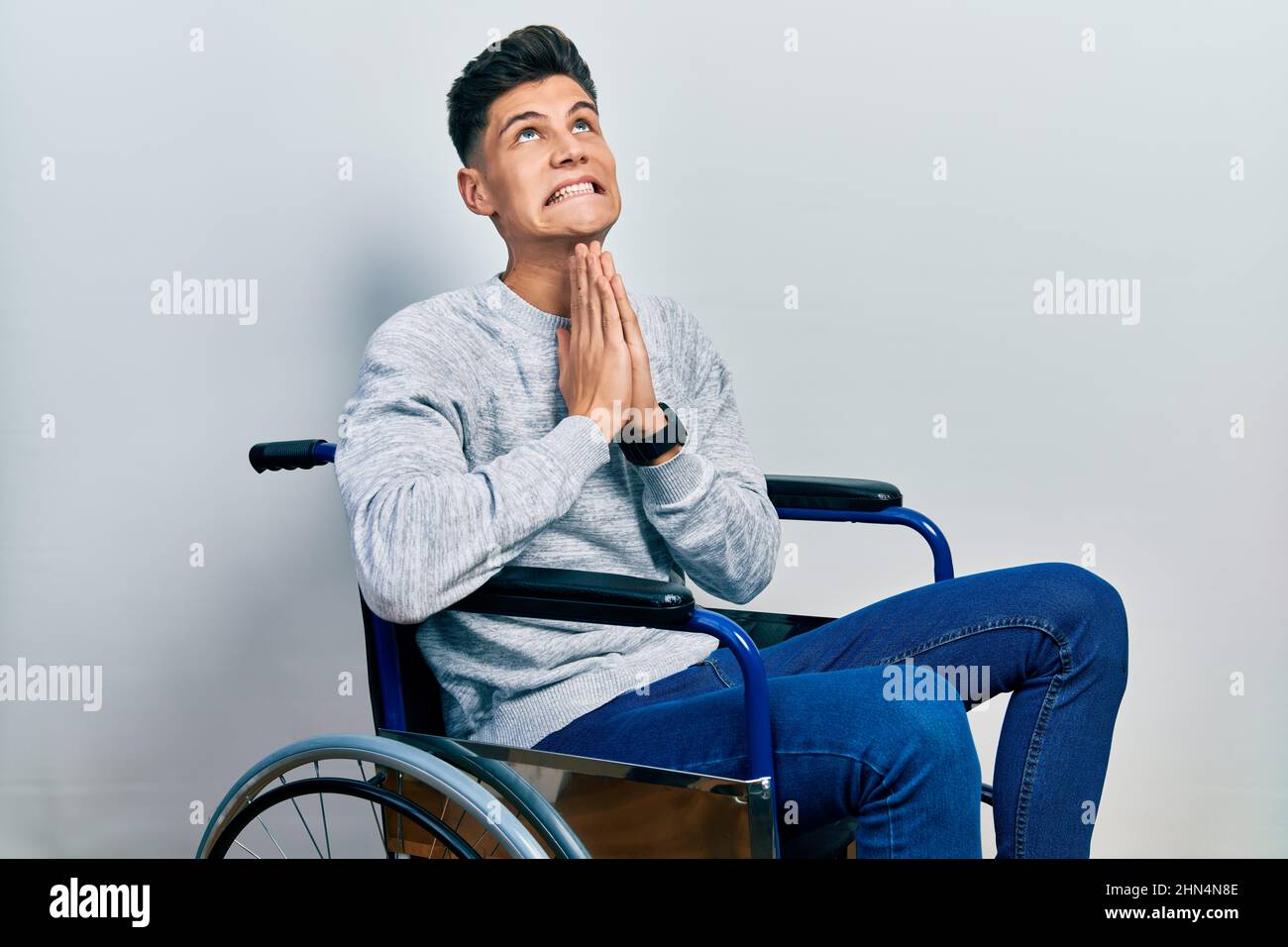 Young hispanic man sitting on wheelchair begging and praying with hands ...