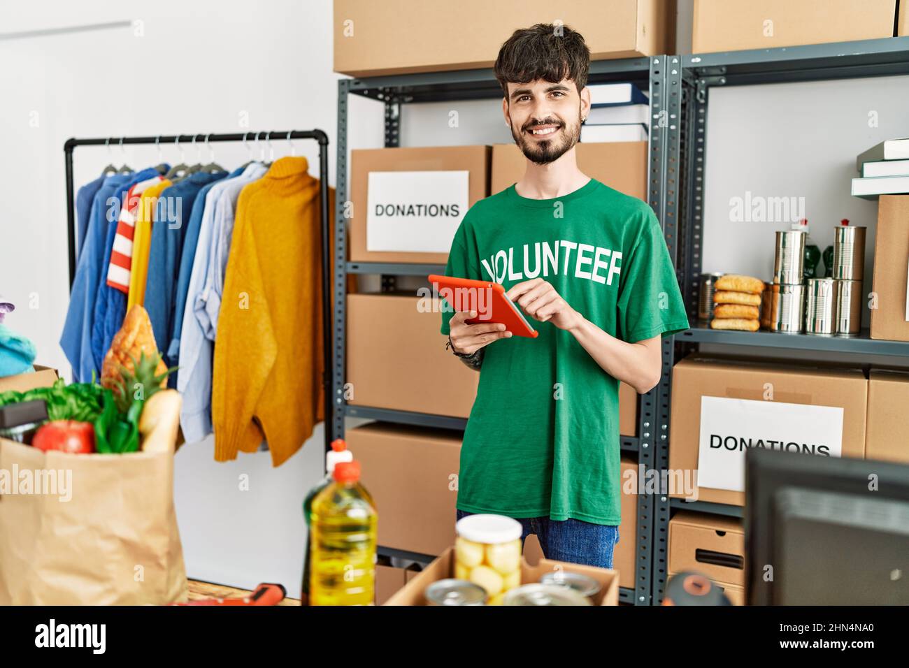 Young hispanic man wearing volunteer uniform using touchpad at charity ...
