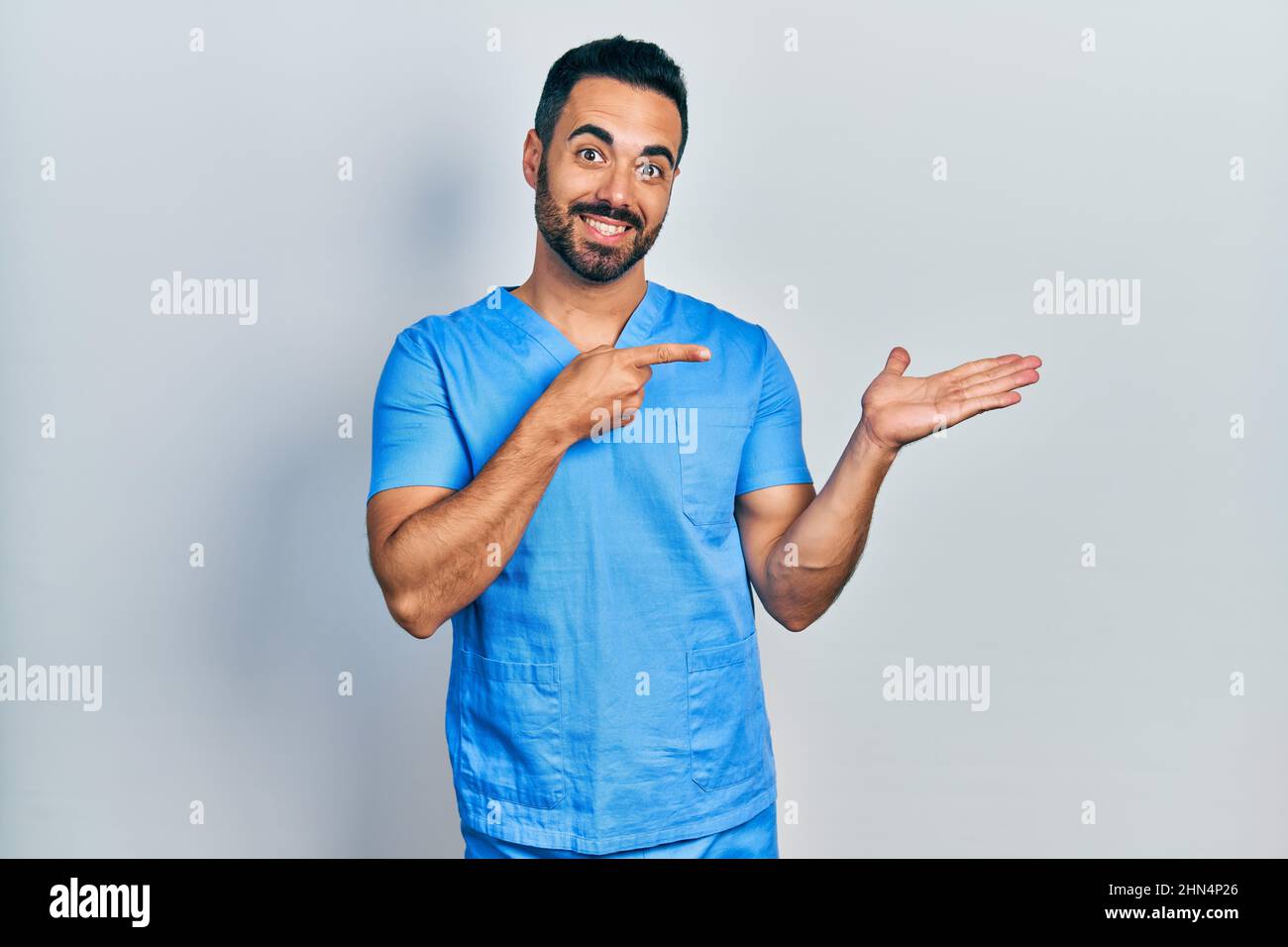 Handsome hispanic man with beard wearing blue male nurse uniform amazed ...