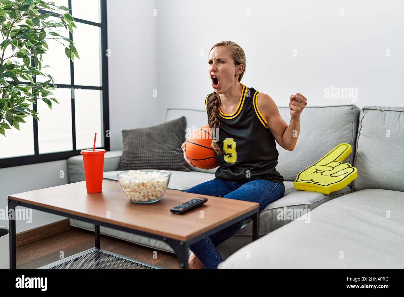 Young blonde woman holding basketball ball cheering tv game annoyed and ...