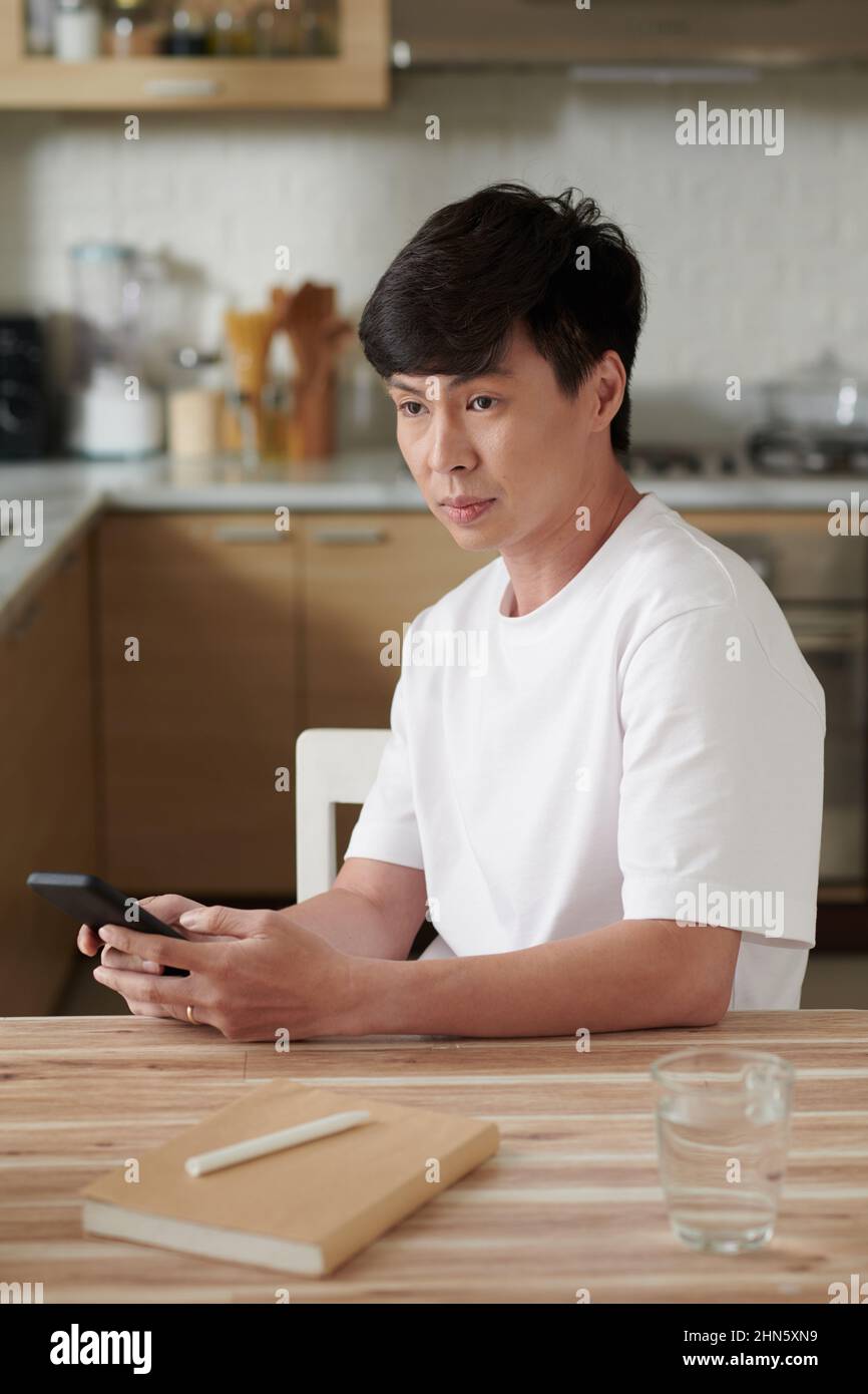Portrait of pensive man sitting at kitchen table with smartphone and ...