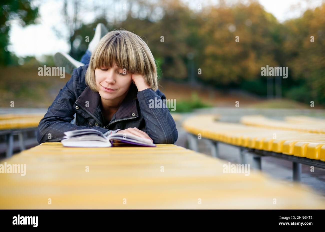 Young woman reading paper book lying on a yellow bench in a public park ...