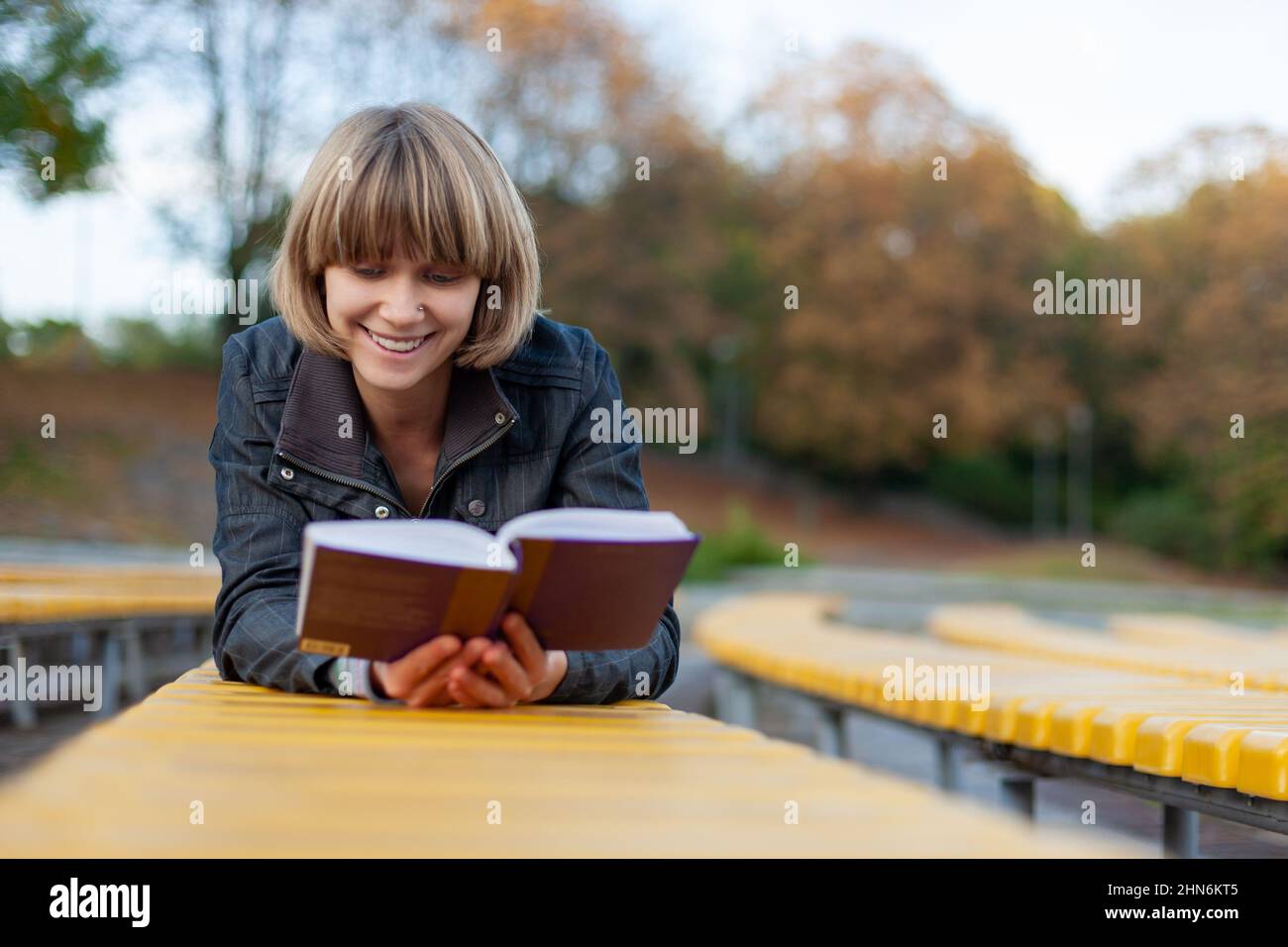 Young woman reading paper book lying on a yellow bench in a public park ...