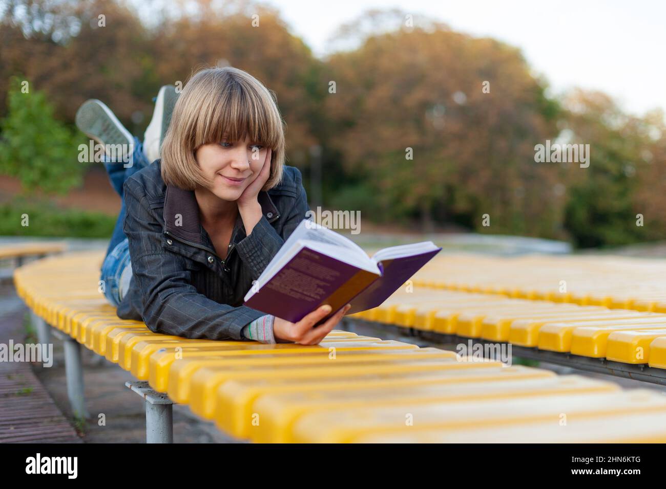 Young woman reading paper book lying on a yellow bench in a public park ...