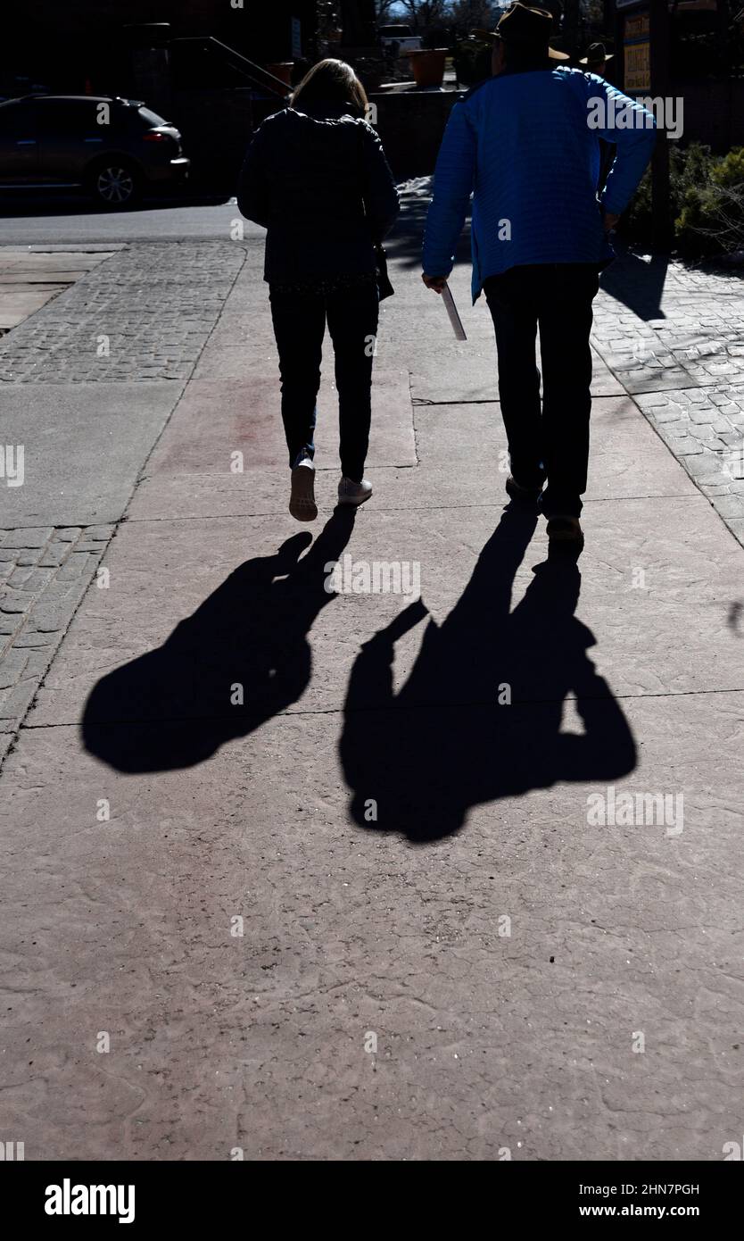 A couple cast long shadows as they walk along a sidewalk in Santa Fe ...