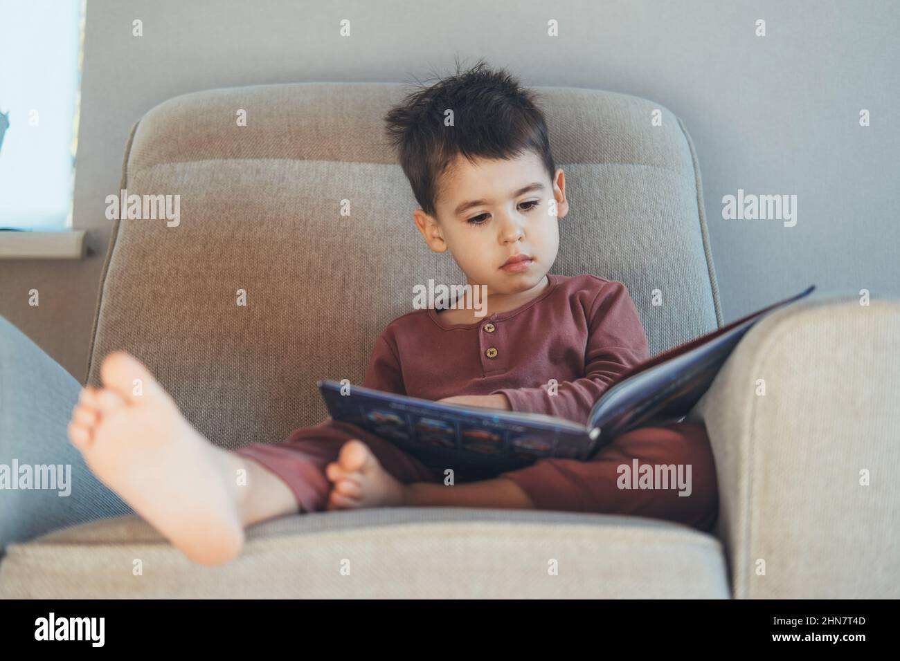 Front view of an little boy reading a book while relaxing on the sofa ...
