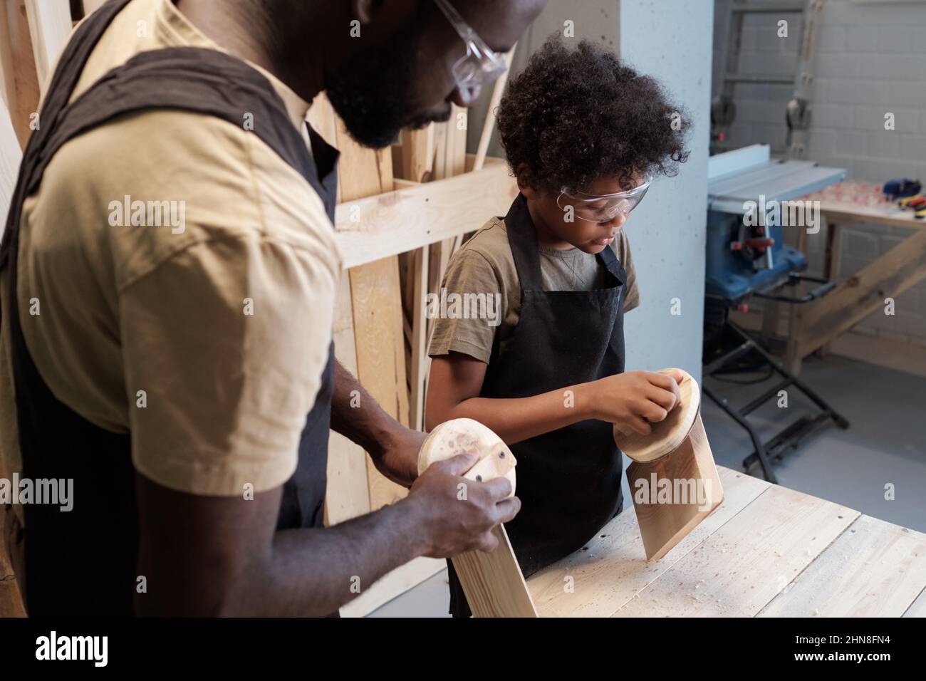 Portrait of black father and son sanding wood in carpentry workshop ...