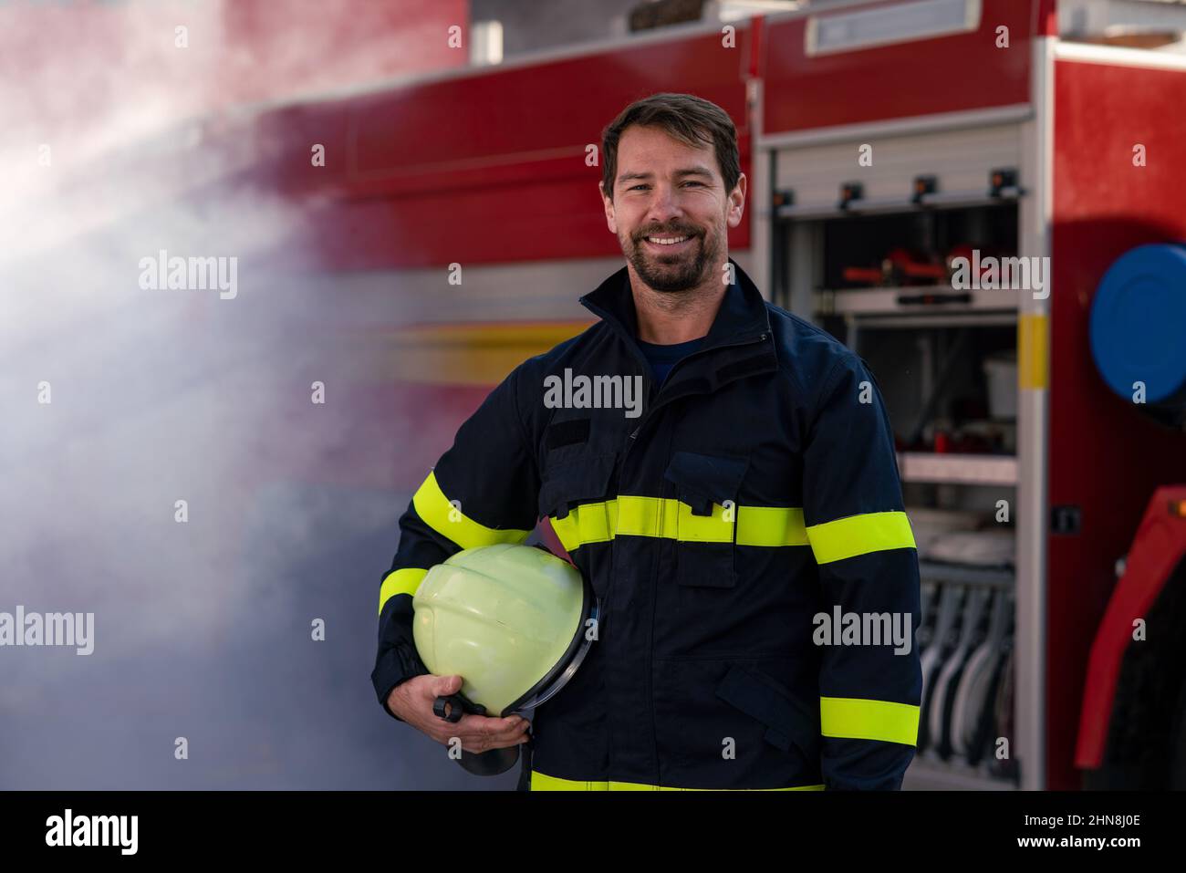 Happy mid-adult firefighter man looking at camera with fire truck and ...