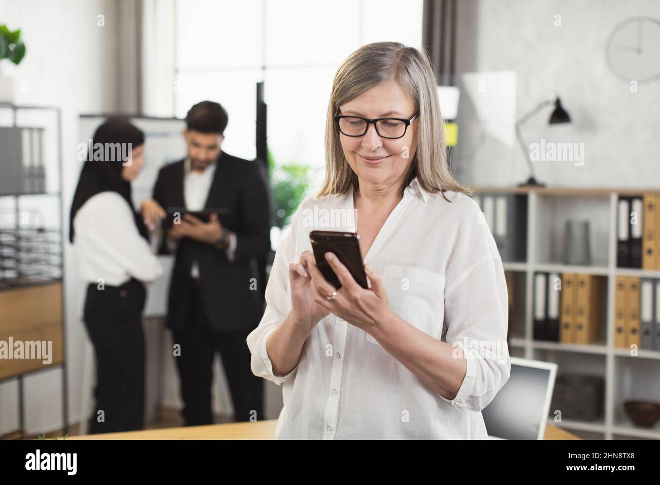 Caucasian business lady with grey hair standing at office and using ...