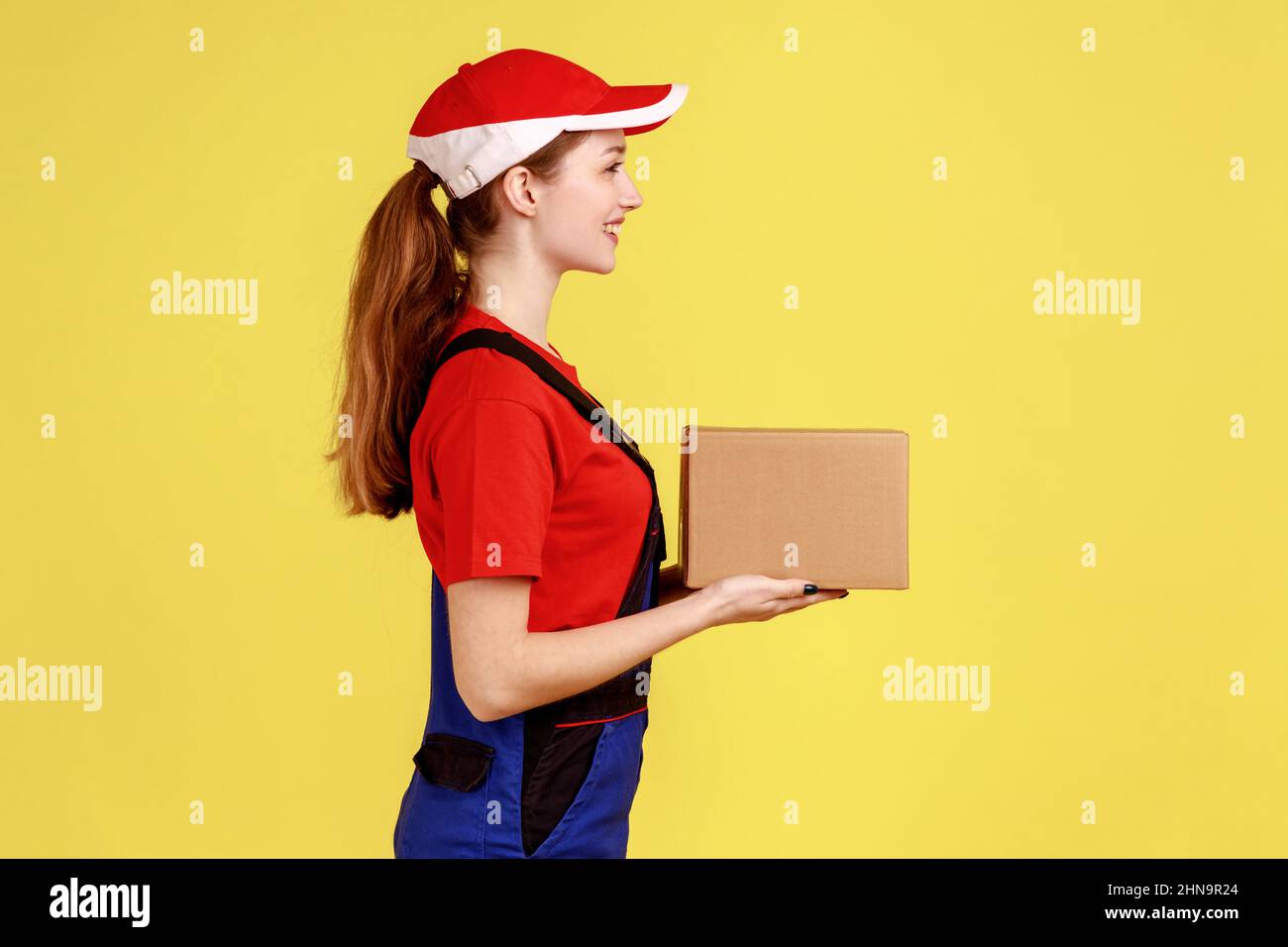 Side view of delighted courier woman standing with cardboard parcel ...