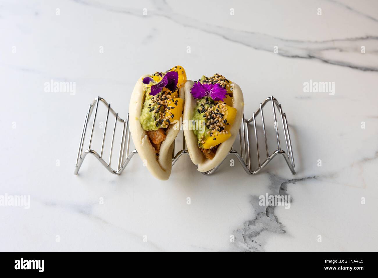 Two white tacos with different foods and purple flower on a stand lined ...