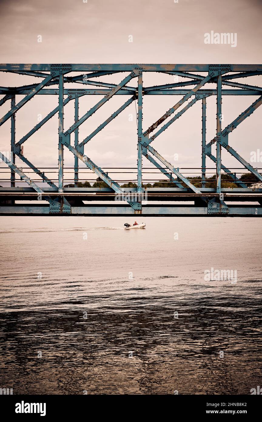 Small motor boat with one occupant sailing under disused box bridge on ...