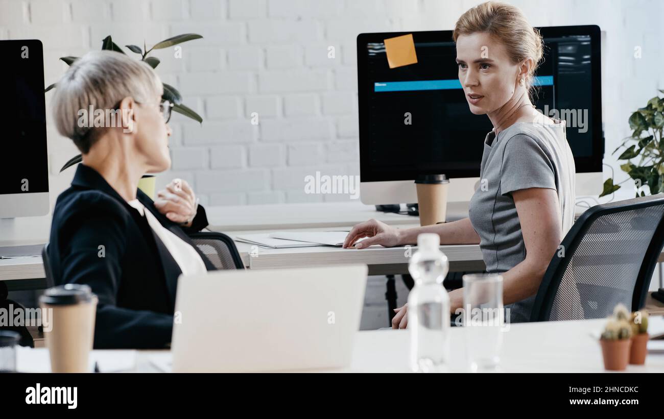 Businesswoman talking to colleague near coffee to go and computers in ...