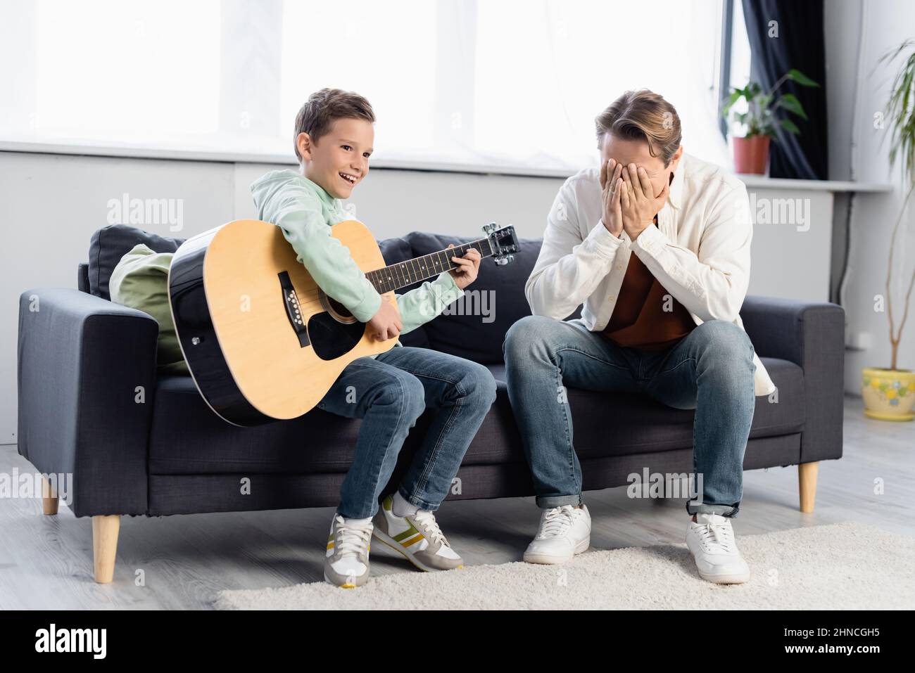 Smiling boy playing acoustic guitar near father covering face on couch ...