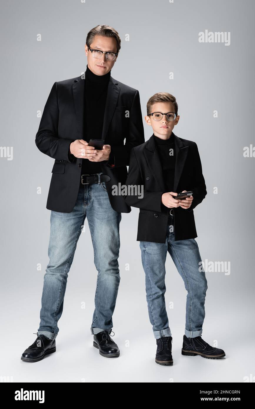 Full length of stylish parent and boy holding mobile phones on grey ...