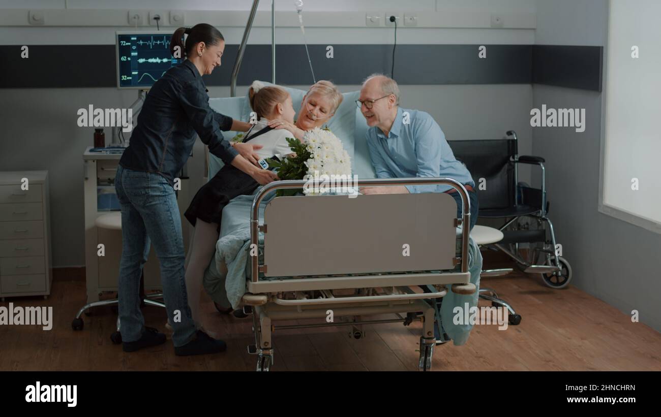 Mother and child visiting sick grandma in hospital ward bed, giving ...