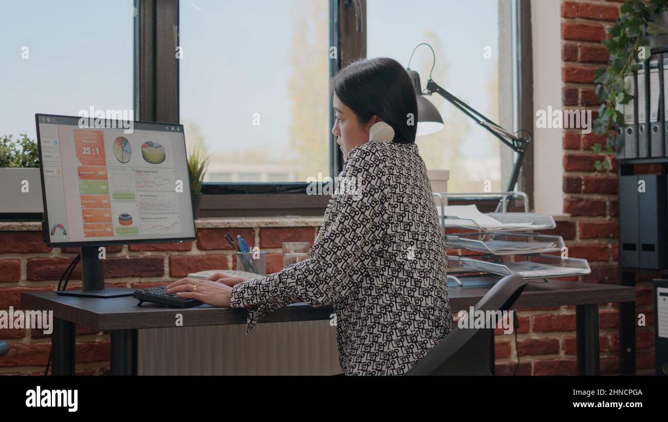 Asian person using office telephone while she works on computer ...