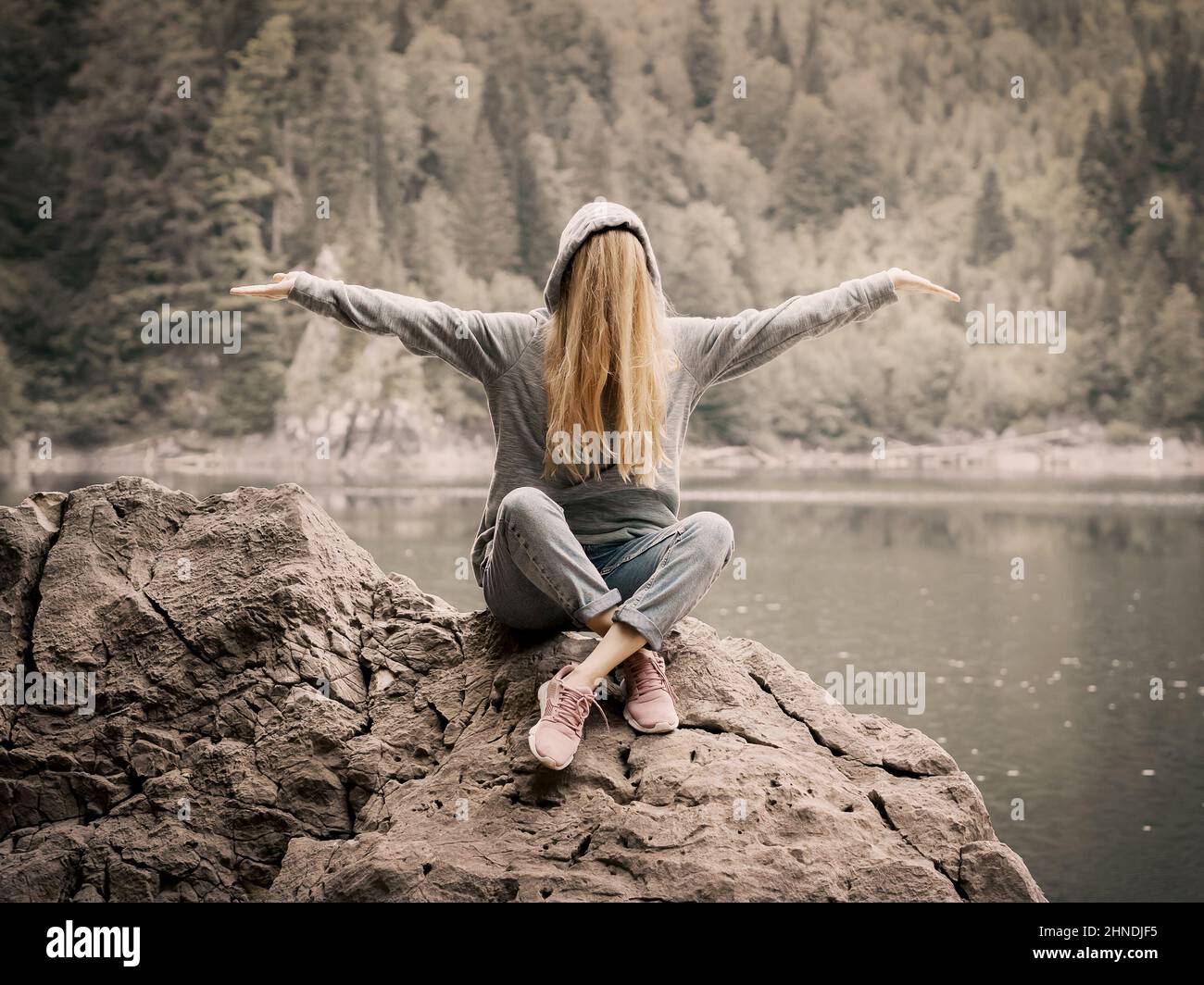 A woman sits on large stone with her arms outstretched and her face ...