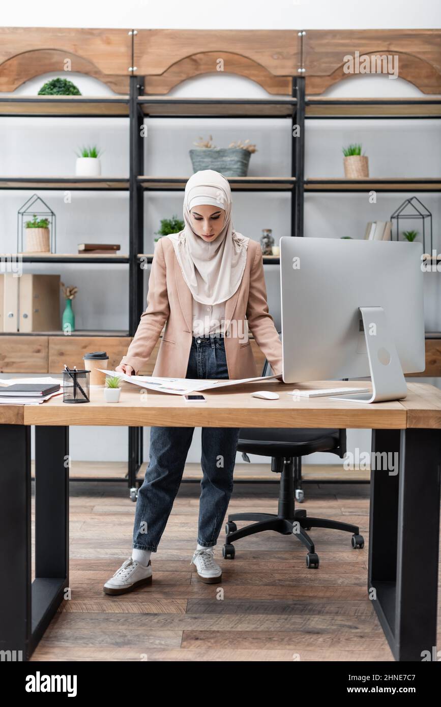 full length view of muslim woman looking at documents near work desk at ...