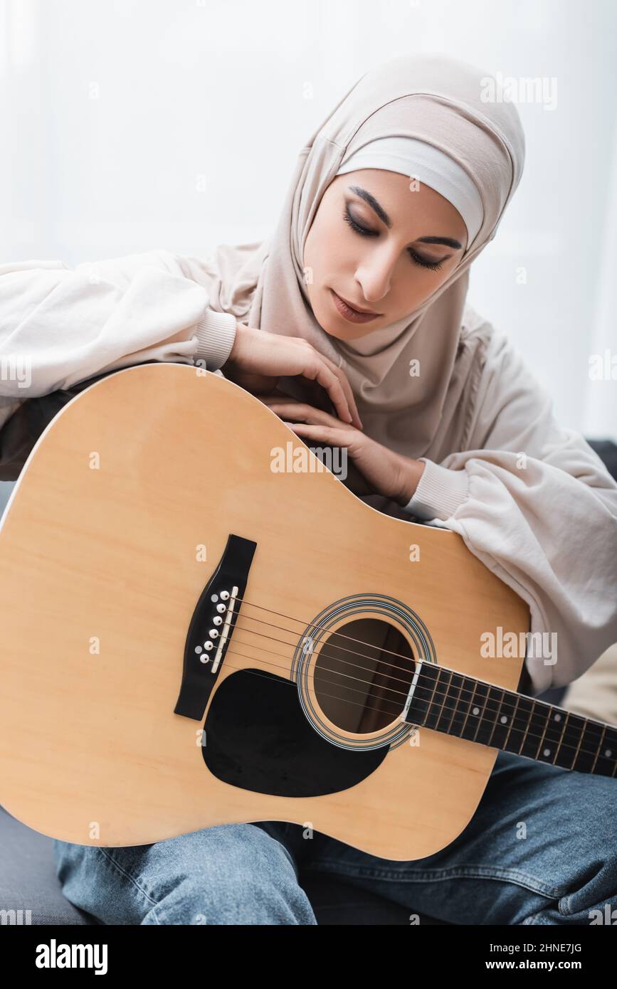young arabian woman in hijab leaning on acoustic guitar while sitting ...
