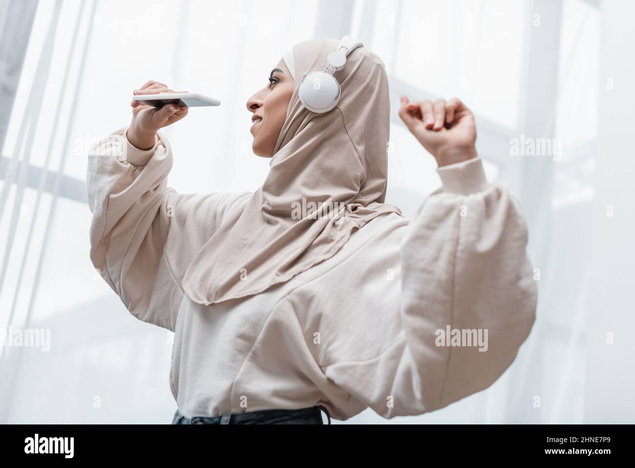 happy arabian woman in hijab and headphones holding smartphone while ...