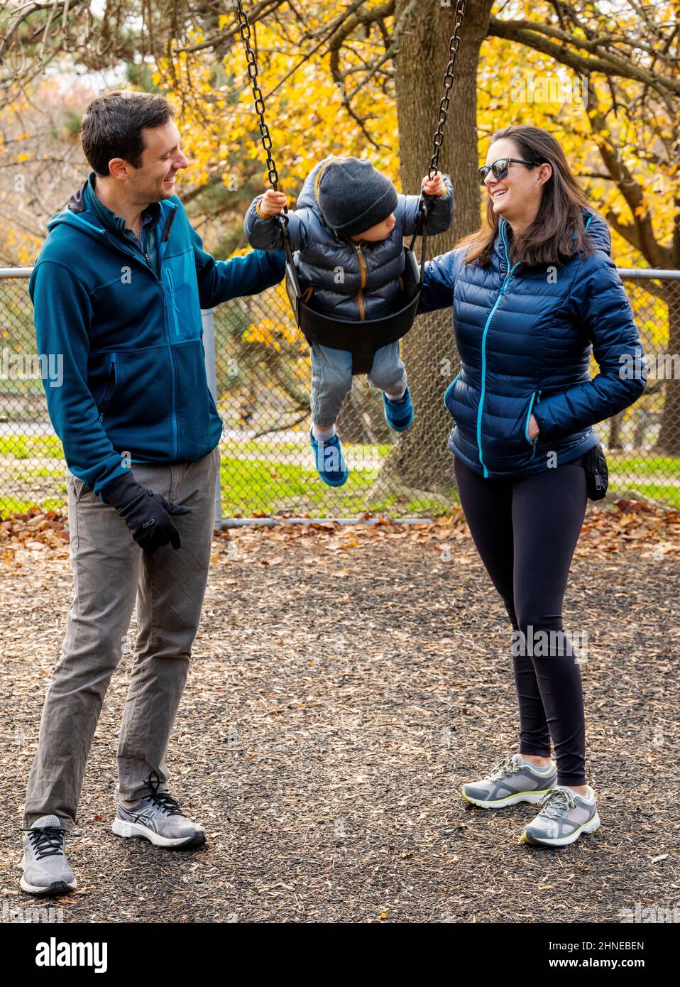 Young parents pushing two-year-old boy on a city playground swing ...