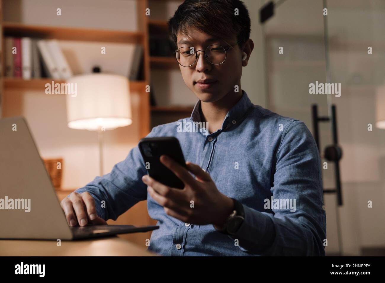 Focused asian guy in earphones using mobile phone while working with ...
