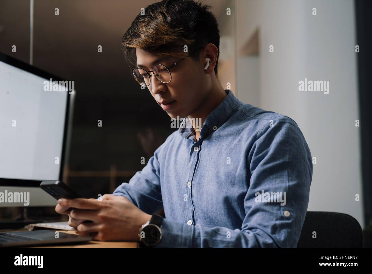 Serious asian guy in earphones using cellphone while working with ...