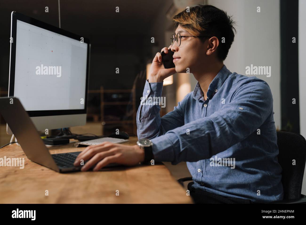Focused asian guy talking on cellphone while working with computers at ...