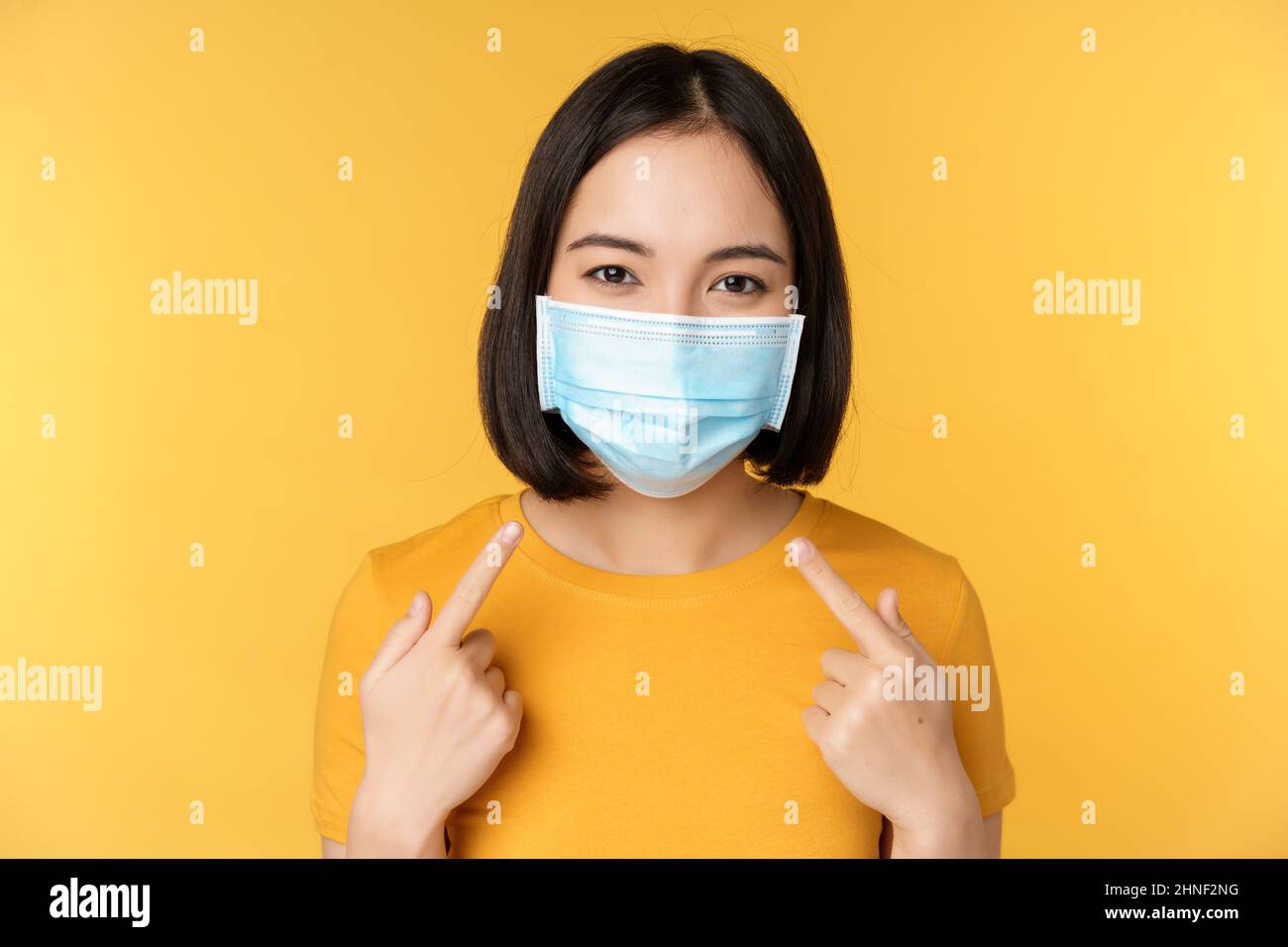 Portrait of smiling asian woman in medical face mask, pointing at her ...
