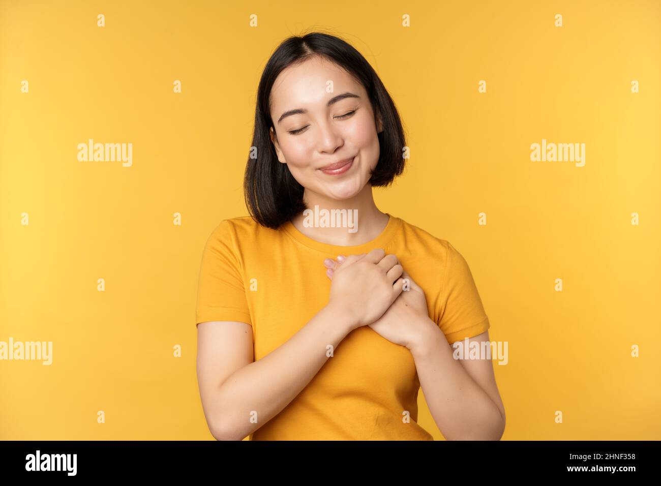 Romantic korean girl, asian woman holding hands on heart, smiling with care and tenderness ...