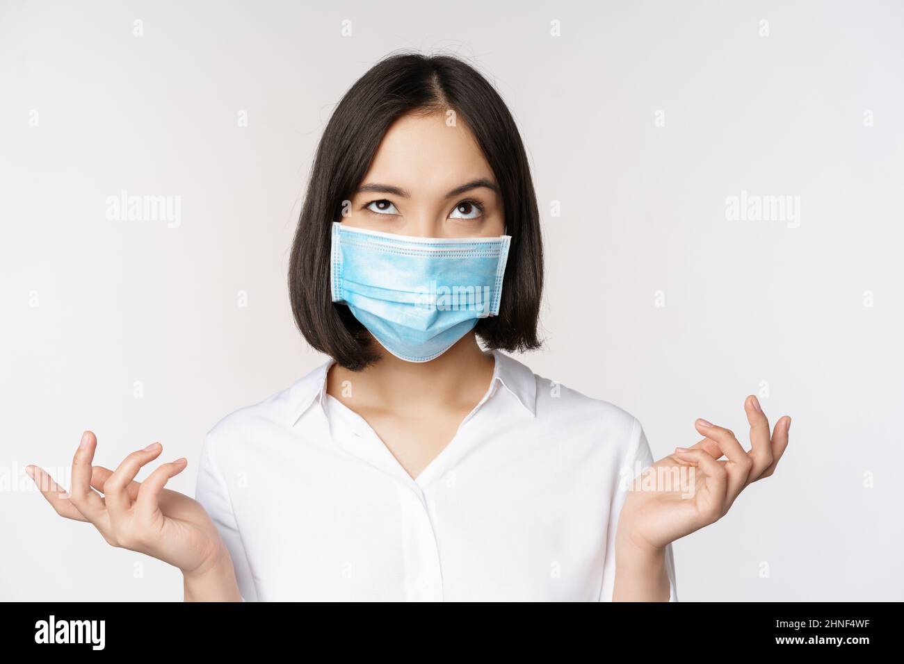 Annoyed asian woman in medical face mask, shrugging and looking up with ...