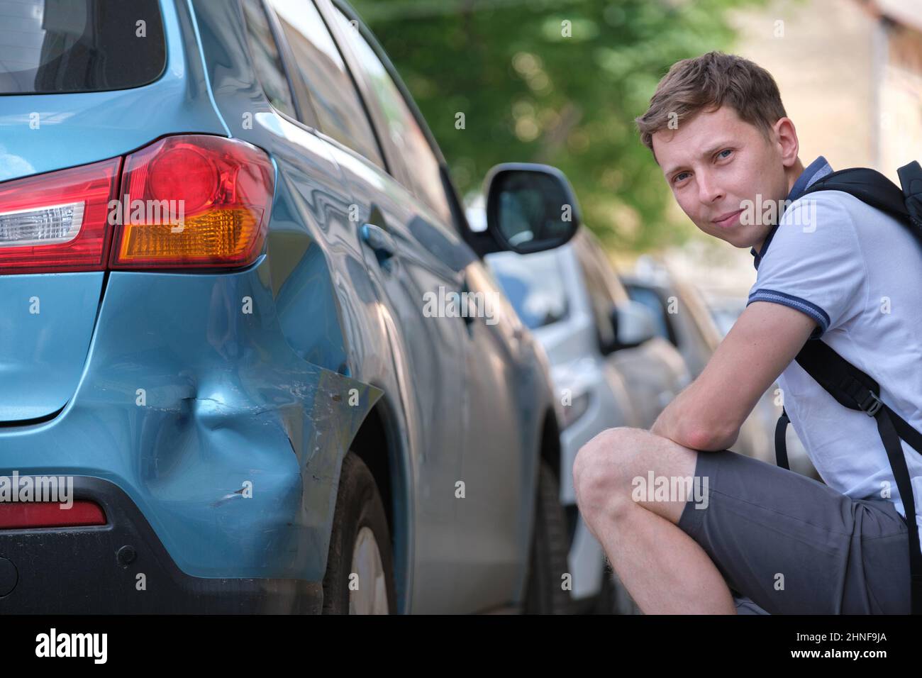 Sad young man driver sitting near his dented car looking shocked on ...