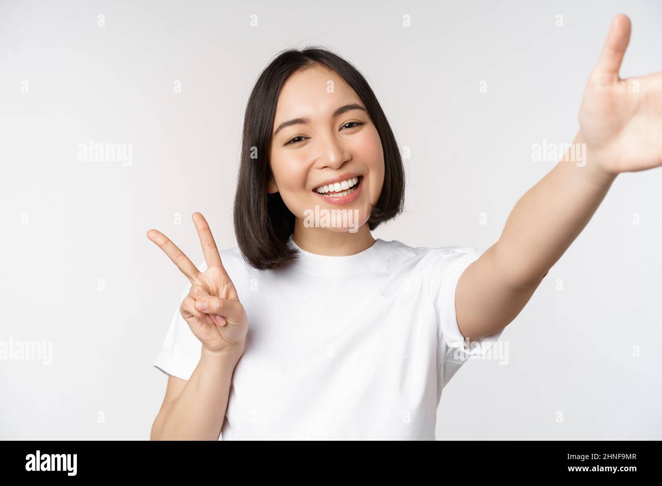 Beautiful young asian woman taking selfie, posing with peace v-sign, smiling happy, take photo ...