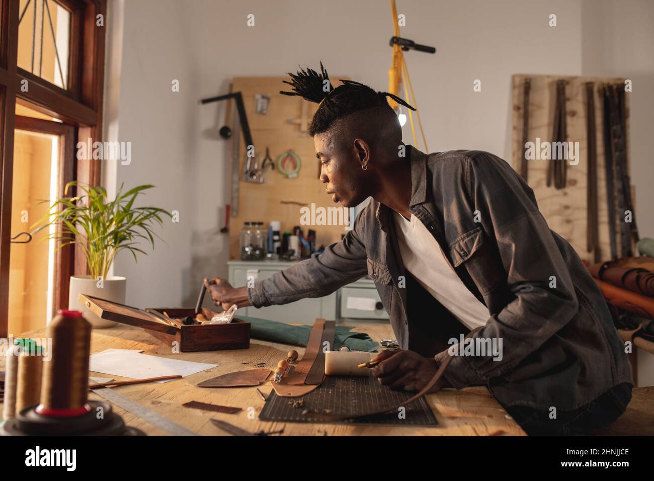 African american young craftsman making belt at workbench in leather ...
