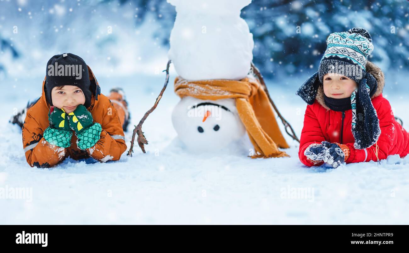Children lying on the snow near a funny snowman. Kids playing outdoors ...
