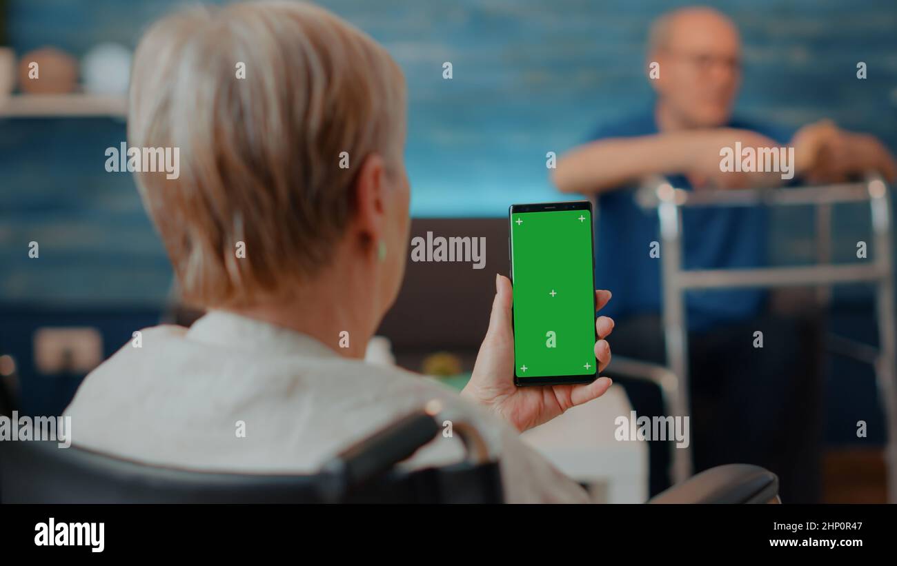 Senior woman in wheelchair holding smartphone with green screen at home ...