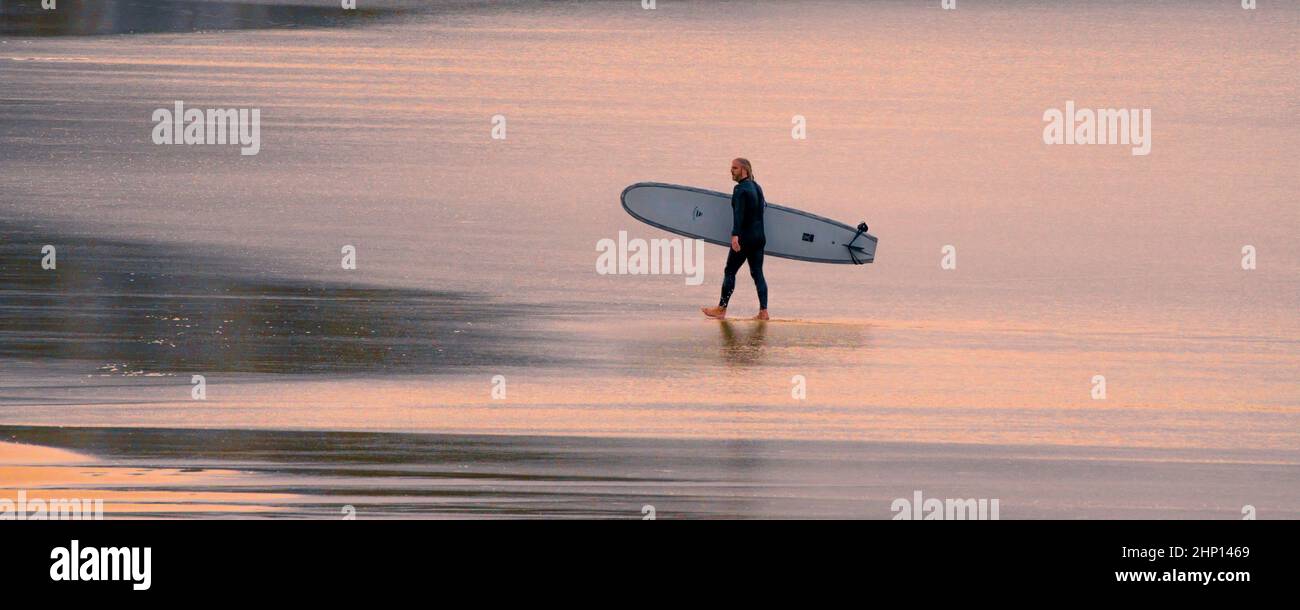 A panoramic image of warm evening light over a surfer carrying his ...
