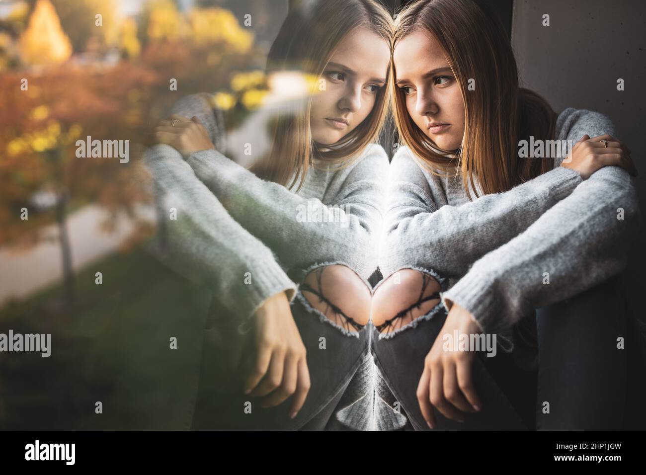 Depressed and anxious young woman sitting by a large window, feeling ...
