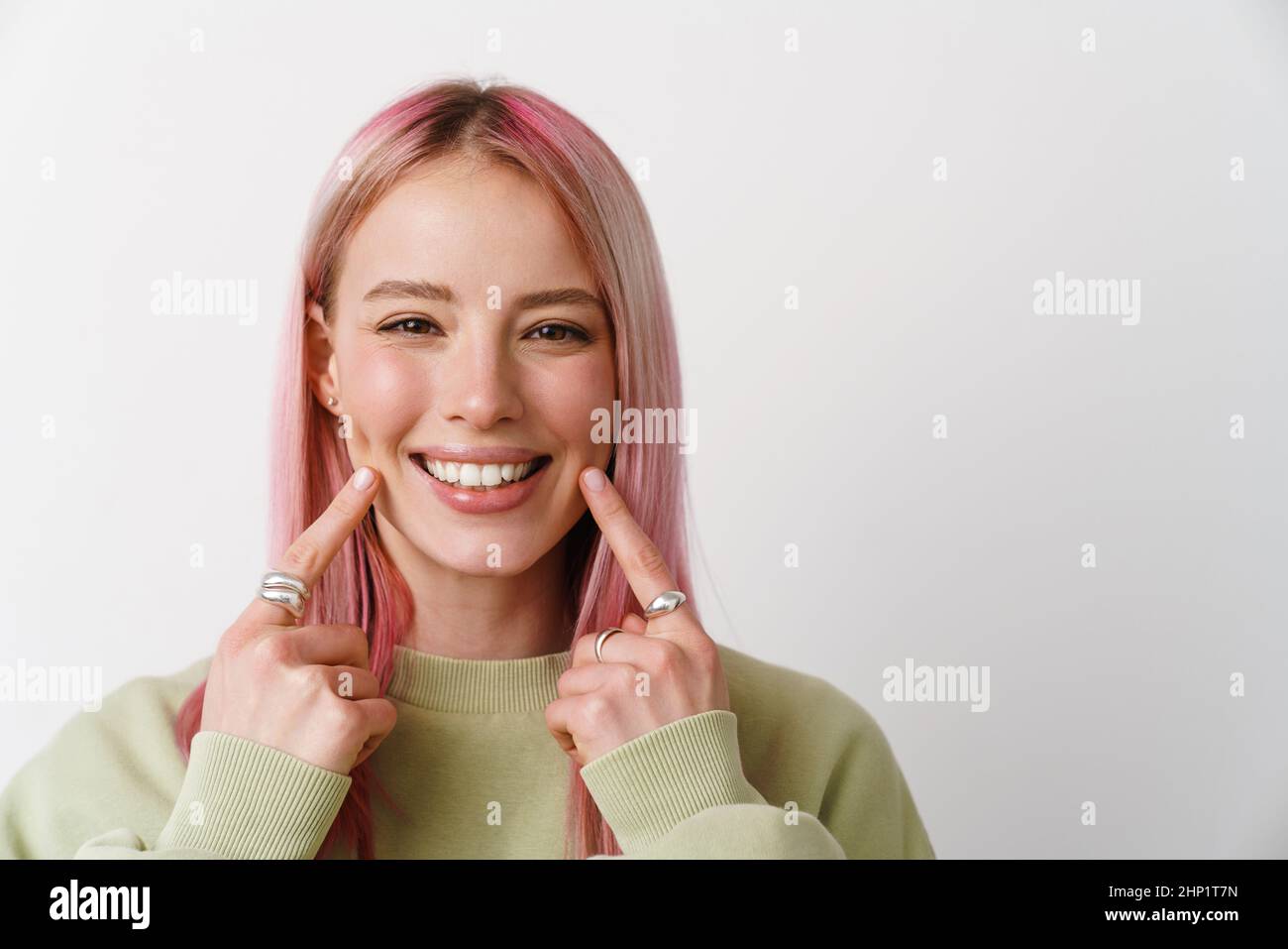 Young smiling woman with pink hair pointing finger at her cheek ...