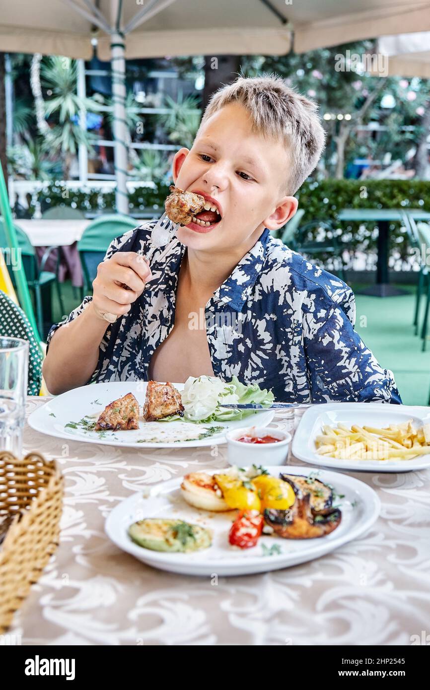 Boy bites and chews piece of meat on fork eating at outdoor table in ...
