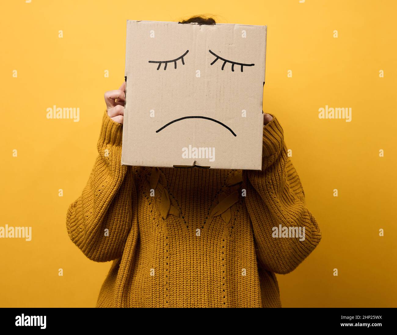 a woman in a brown sweater with a box on her head stands on a yellow ...