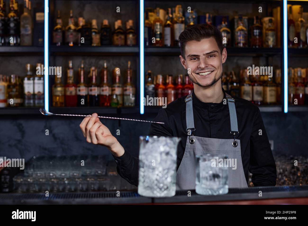 smiling barman in apron stirring ice cubes in cocktail glass with spoon ...