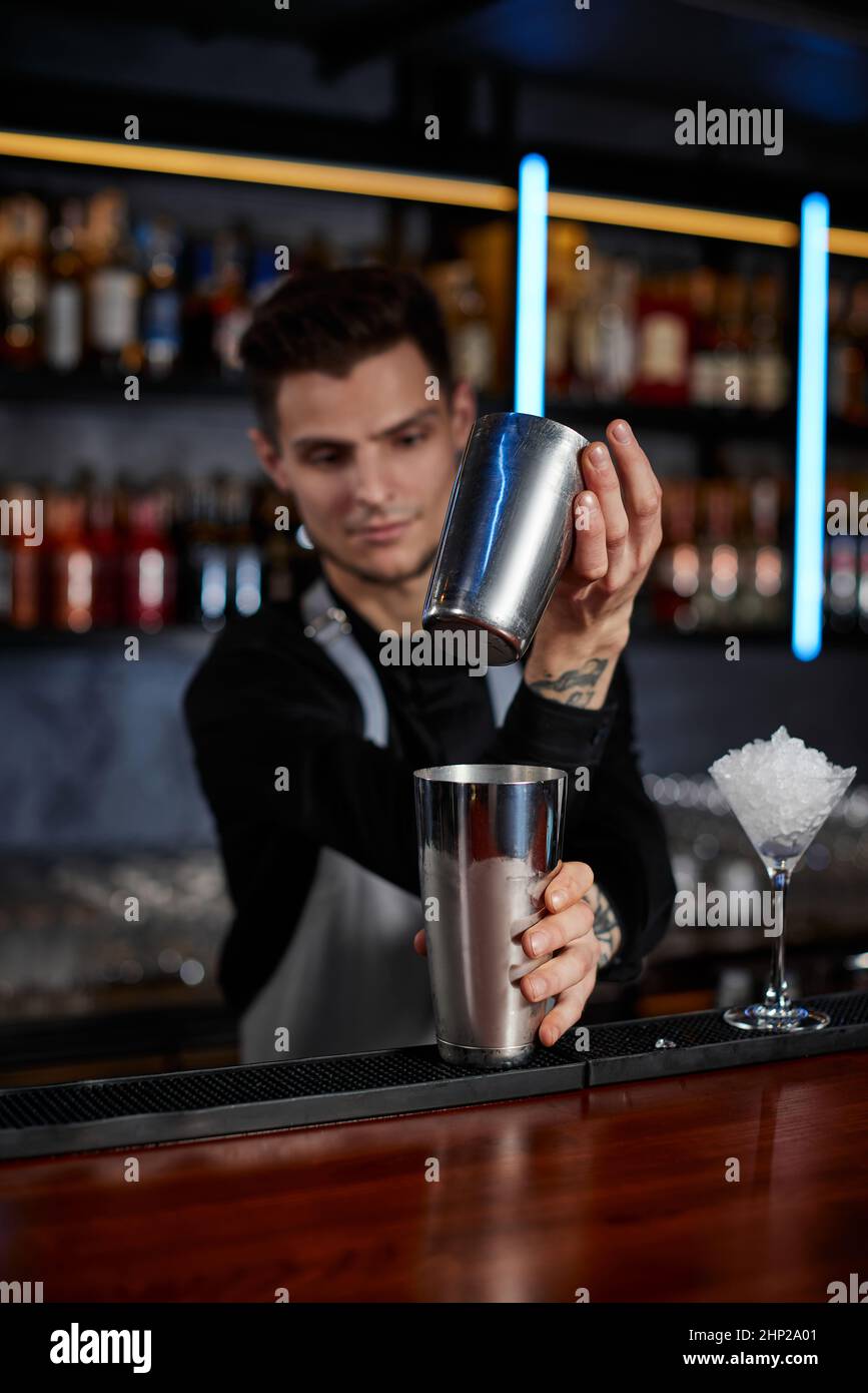 handsome barman is making cocktail with steel shaker at the bar counter ...