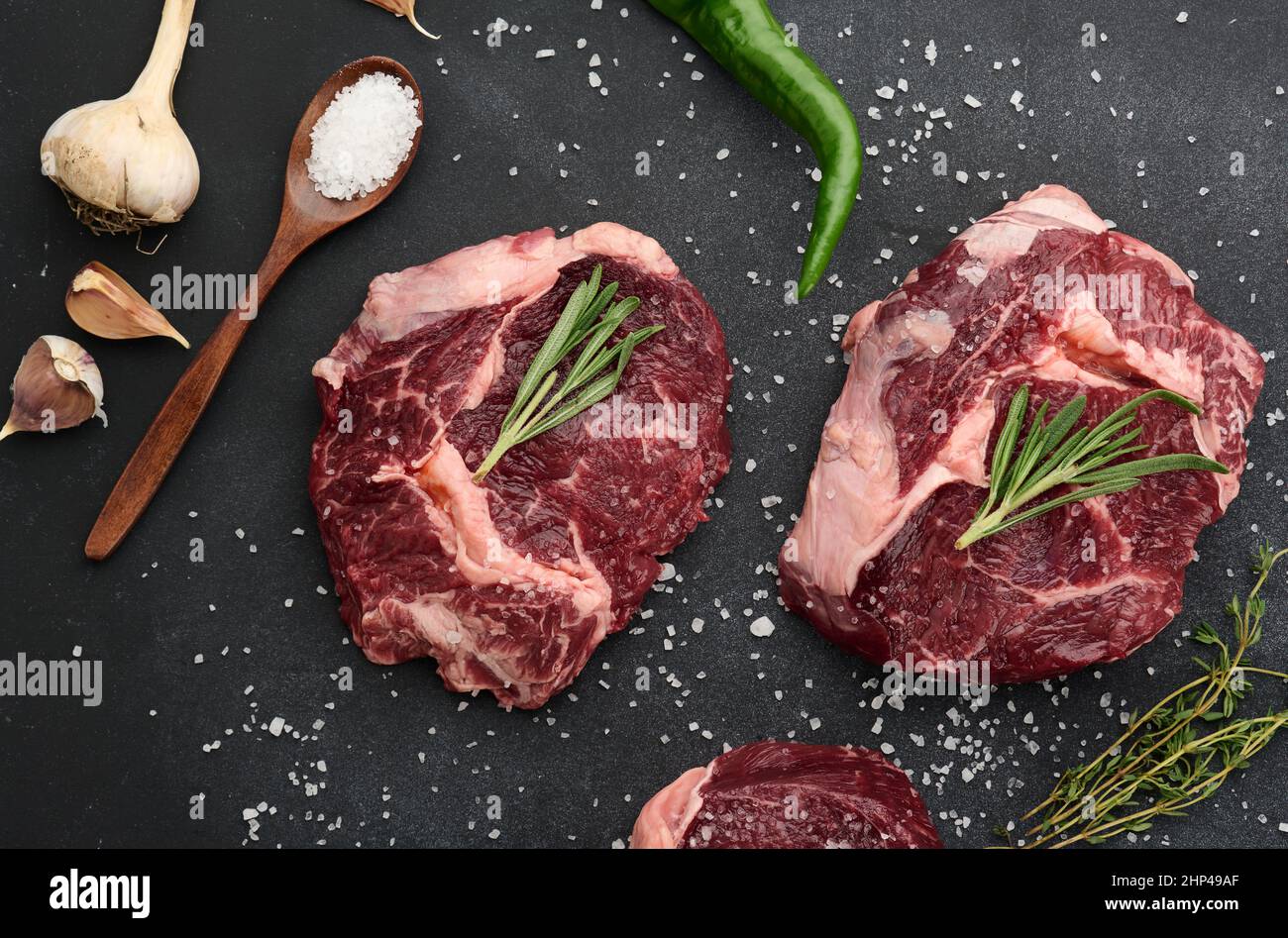 raw piece of beef ribeye with rosemary, thyme on a black table, top ...