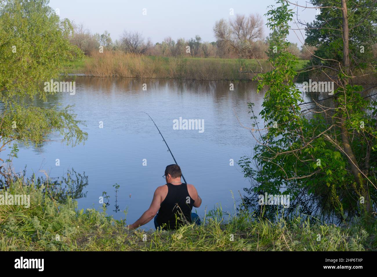 Fisherman sits with fishing rod on bank of the river. View from the ...