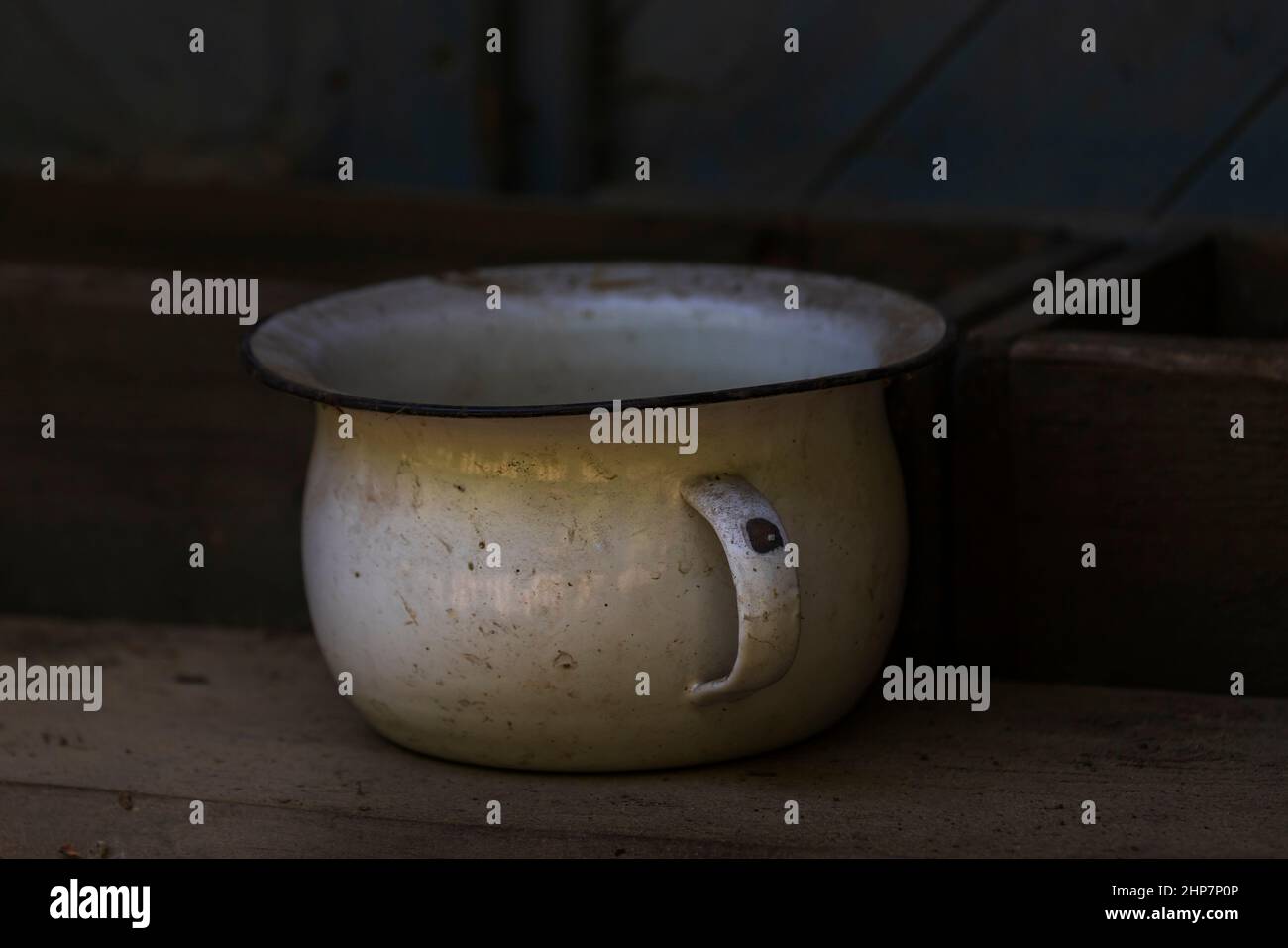 An tin old white battered potty stands on a wooden shelf. The ...
